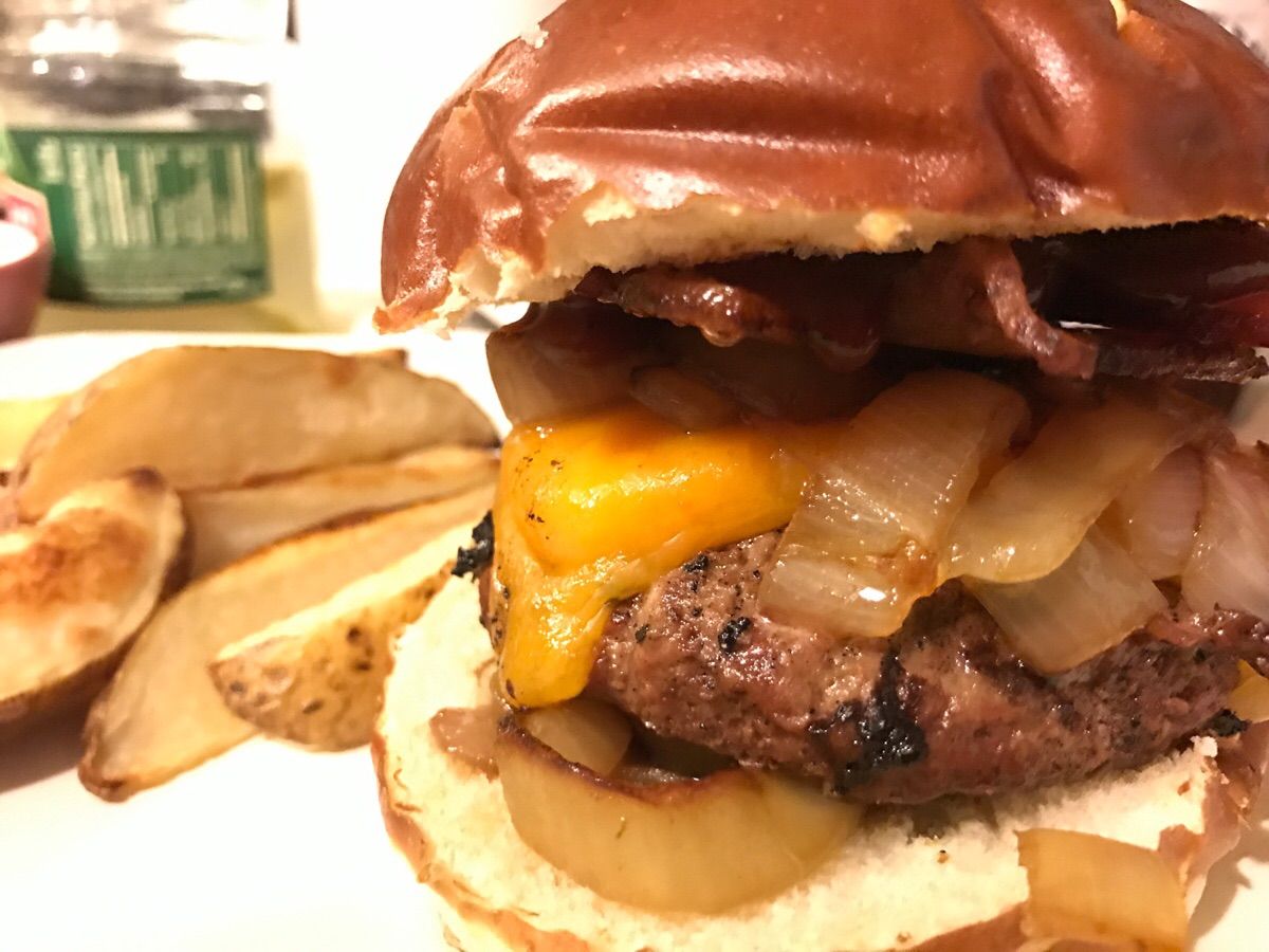 Burger with fried onion, thick cut bacon, cheddar and bbq on a pretzel roll with potato wedges ...